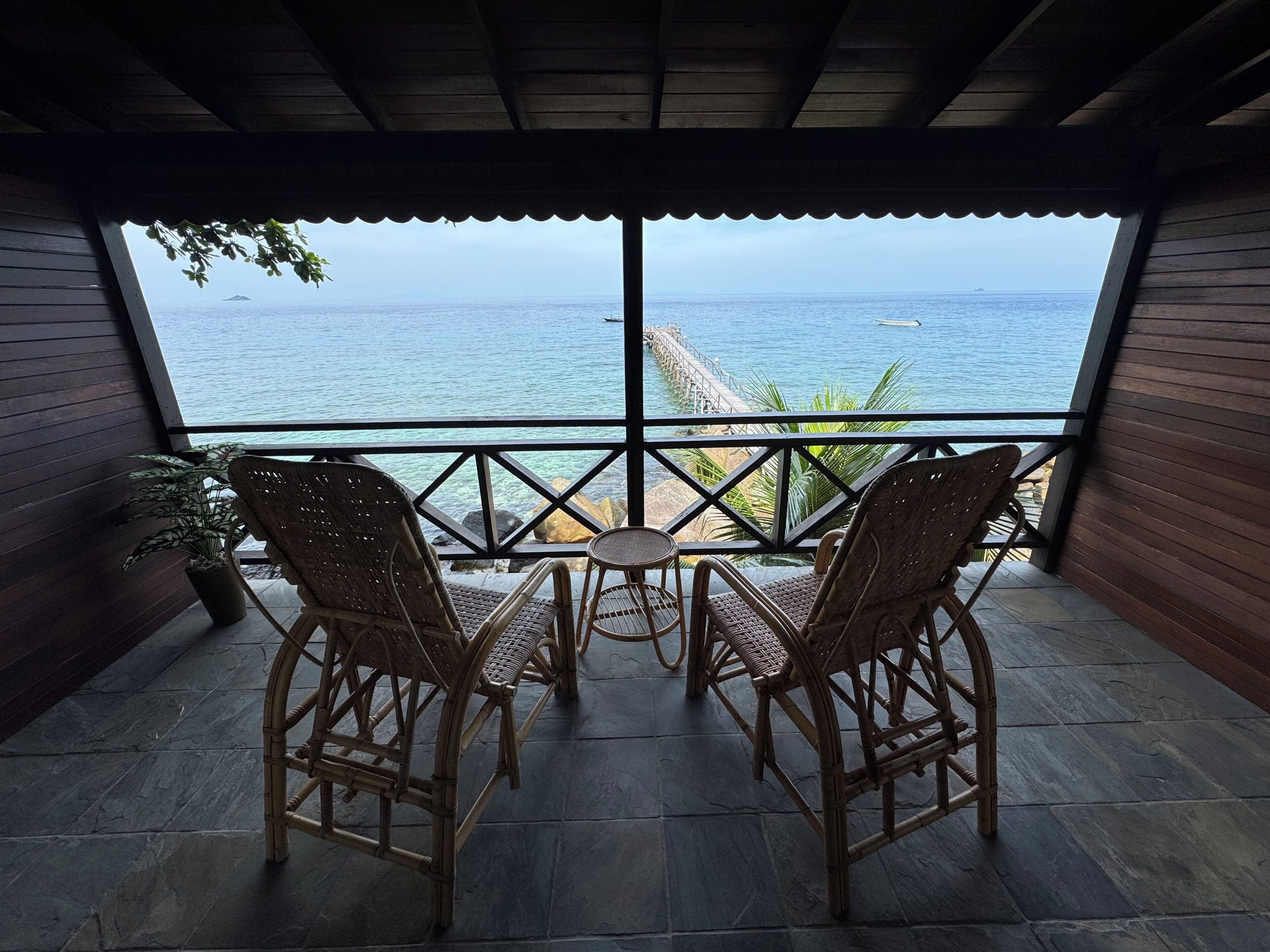 Uma área de estar coberta ao ar livre, com duas cadeiras de vime e uma pequena mesa, com vista para o mar.