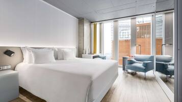 Quarto de hotel moderno com uma cama grande, uma mesinha de cabeceira, um espaço de estar com cadeiras e vista através de uma porta de vidro.