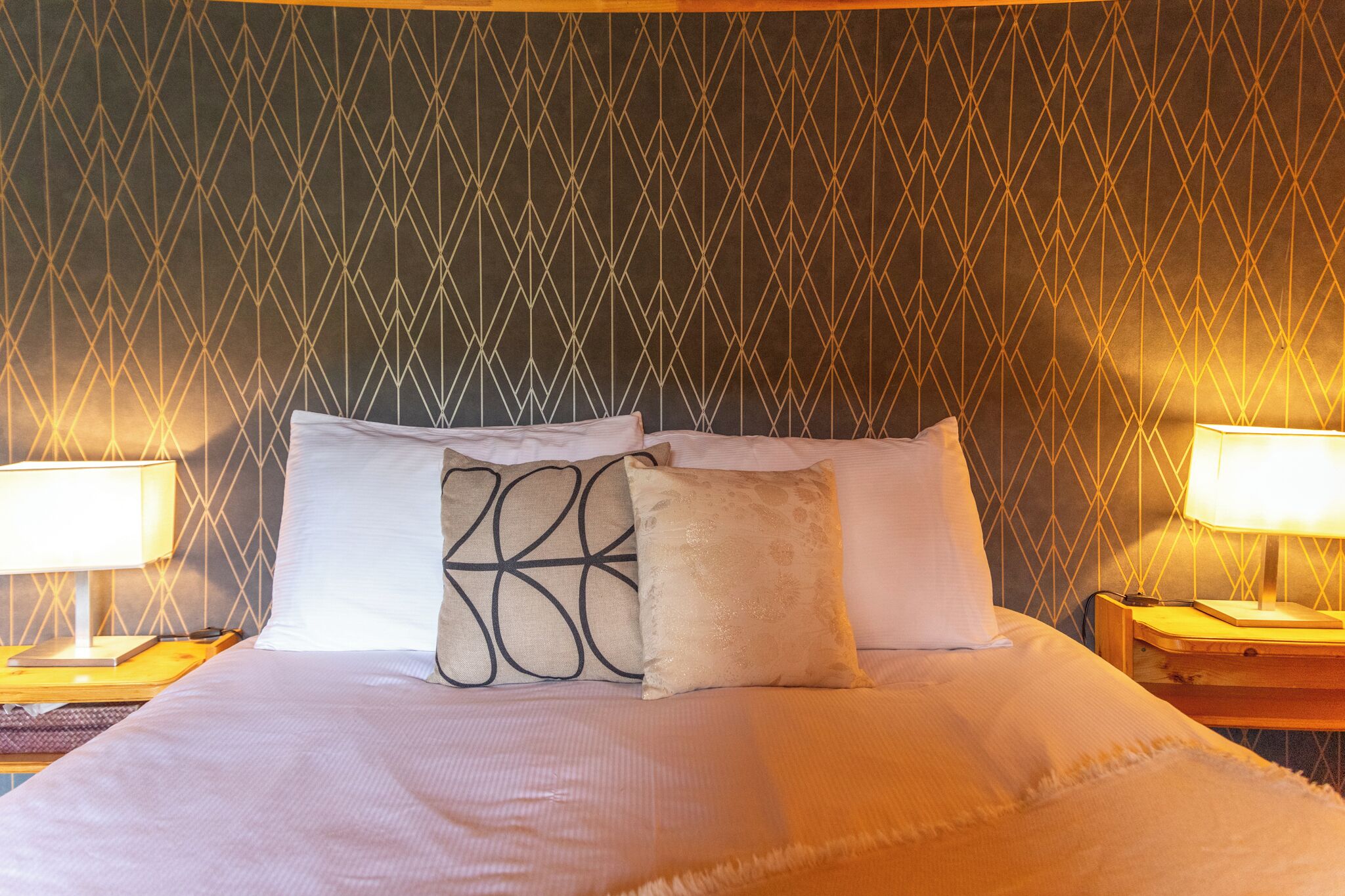 Cama bem arrumada com lençóis brancos e duas almofadas decorativas, mesa de cabeceira com abajur, e parede com padrão atrás da cama.
