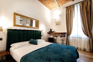 Quarto de hotel com cabeceira estofada verde, um espelho grande, cama com roupa de cama branca e colcha estampada azul, escrivaninha com cadeira e janela com cortinas bege.