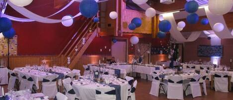 Salão de banquetes com mesas redondas preparadas para um evento formal, com toalhas brancas e fitas azuis.