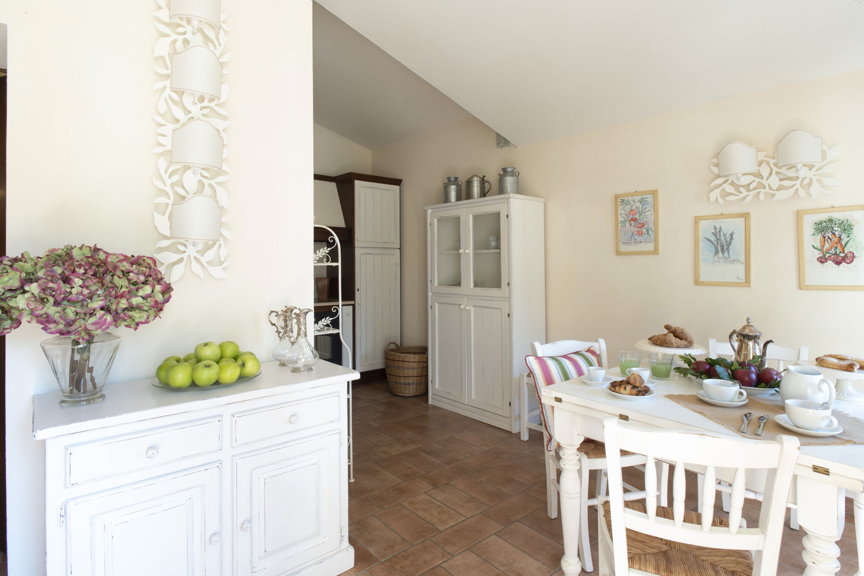 Uma área de refeições em tons de branco, com uma mesa posta para o café da manhã, um armário com portas de vidro e um luminário fixado na parede.