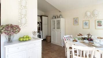 Uma área de refeições em tons de branco, com uma mesa posta para o café da manhã, um armário com portas de vidro e um luminário fixado na parede.