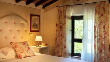 Quarto de hotel com cama grande, cabeceira com botões, mesa de cabeceira com abajur, quadro na parede e cortinas florais.
