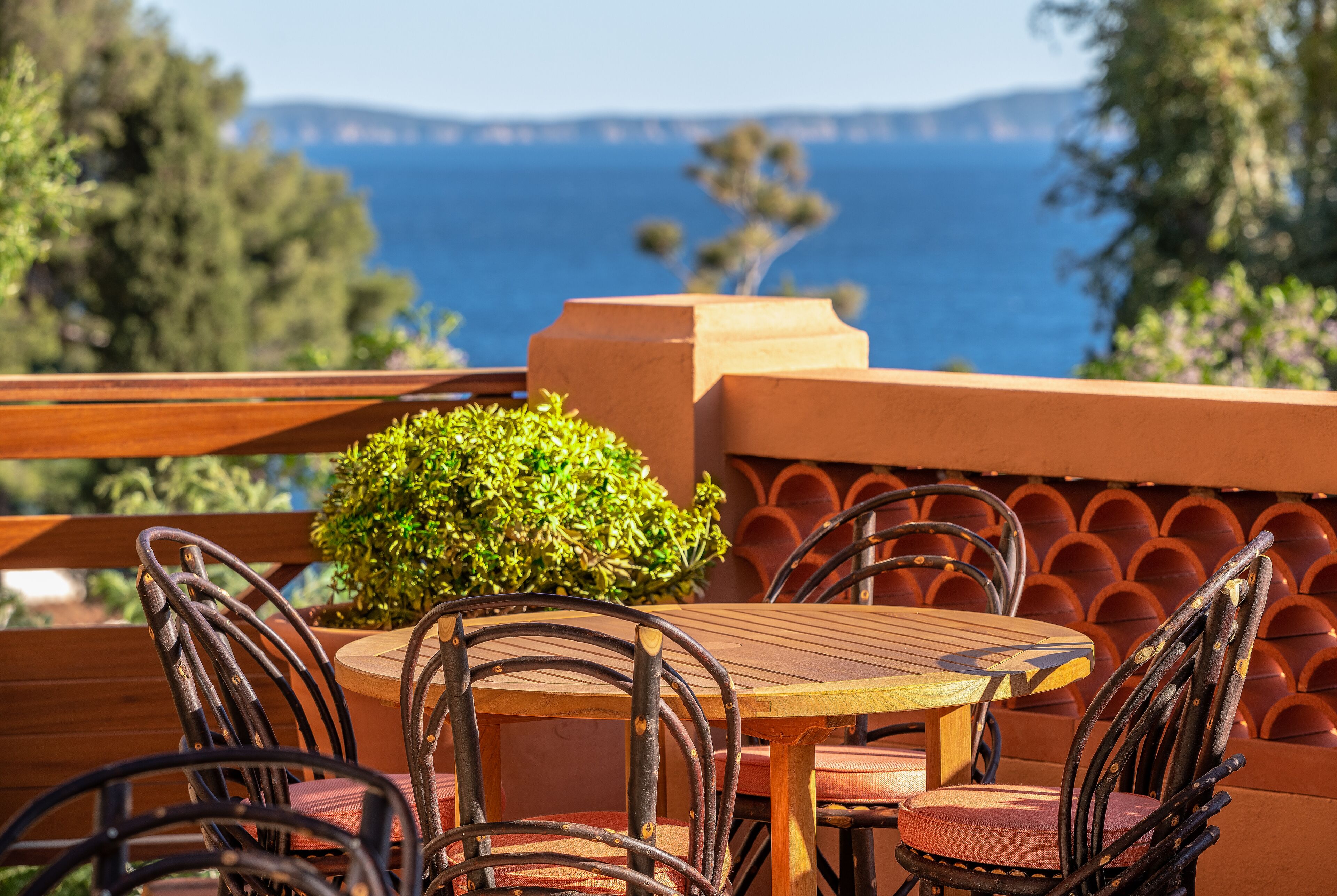 Um terraço com mesa e cadeiras de madeira, um vaso de planta e vista para o mar.