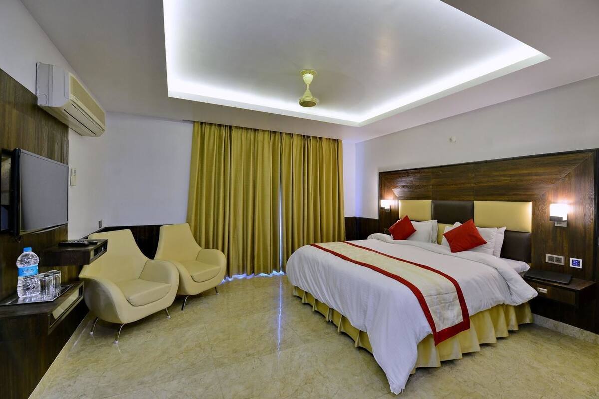 Quarto de hotel com uma cama, duas poltronas, uma escrivaninha com uma garrafa de água, uma televisão e uma luminária no teto.