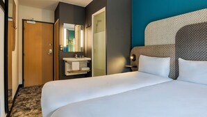 Quarto de hotel com uma cama grande, banheiro com pia e espelho, e porta que dá para outro cômodo.