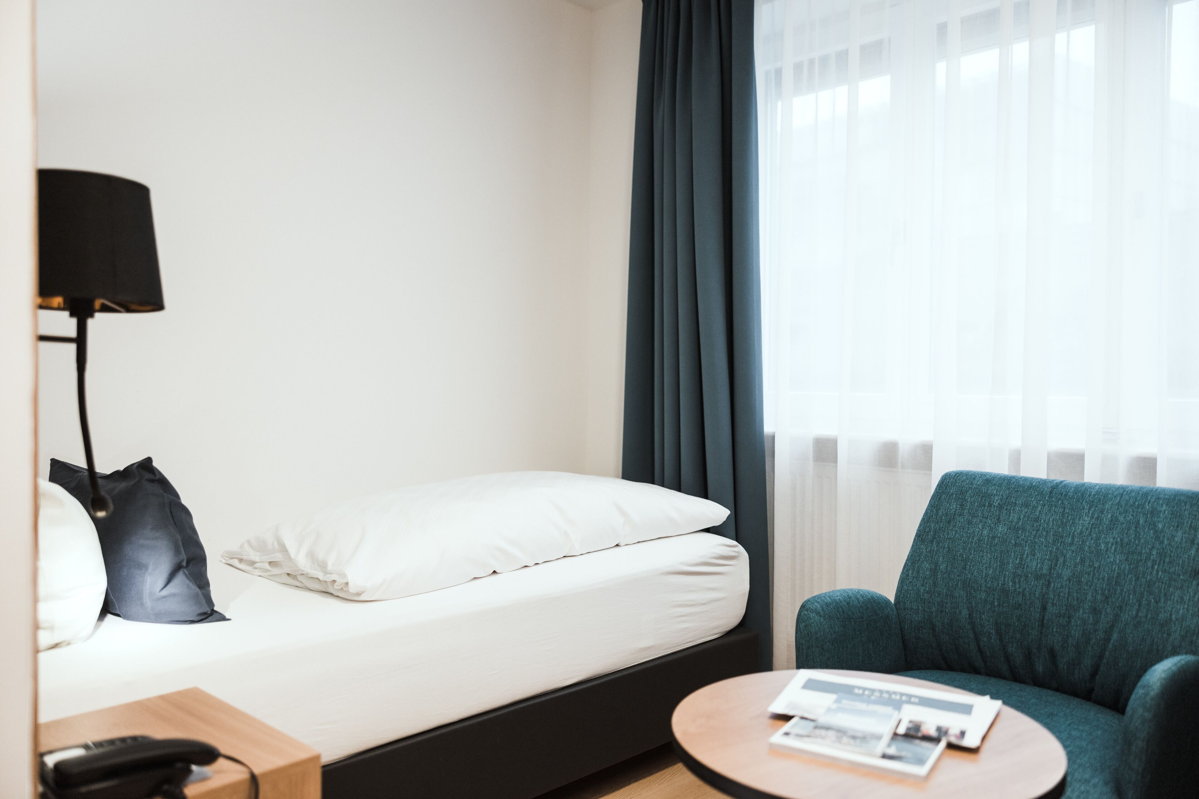 Quarto de hotel com cama, uma cadeira, abajur e mesa com uma revista.