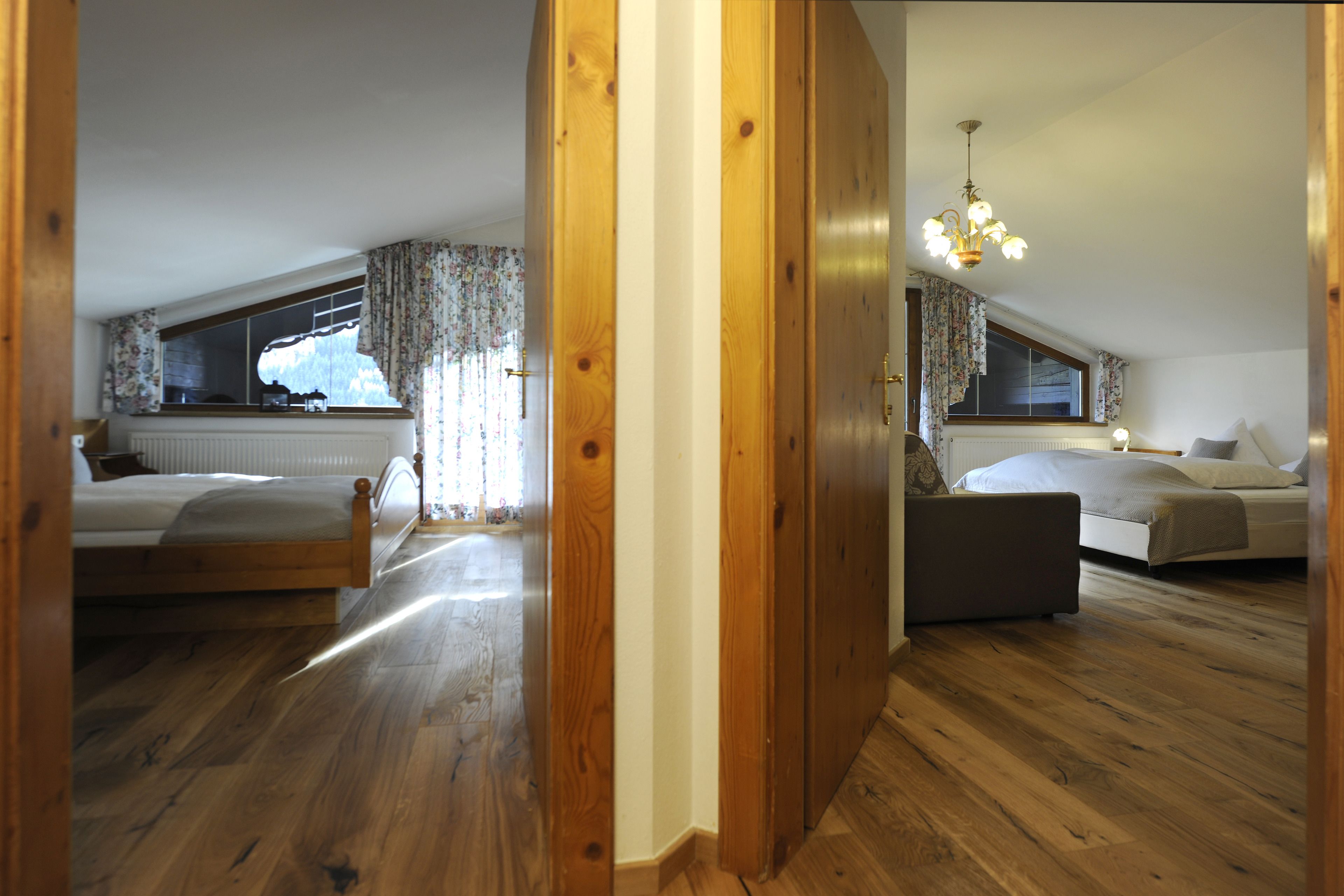 Quarto com teto inclinado, piso de madeira, duas camas, janela com cortinas e um lustre.