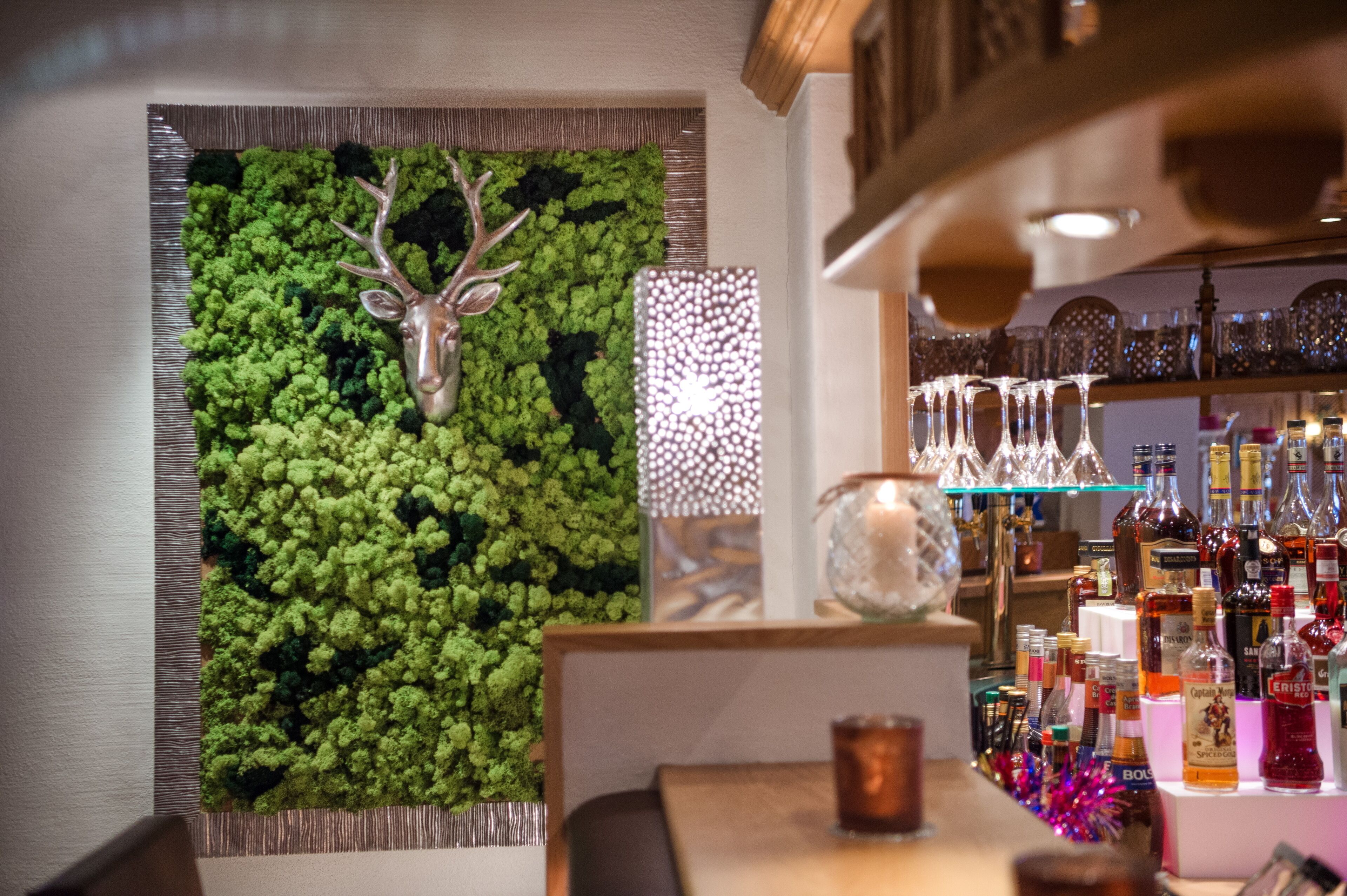 Uma área de bar com parede coberta de musgo verde, decoração de cabeça de veado e diversos frascos de bebidas.