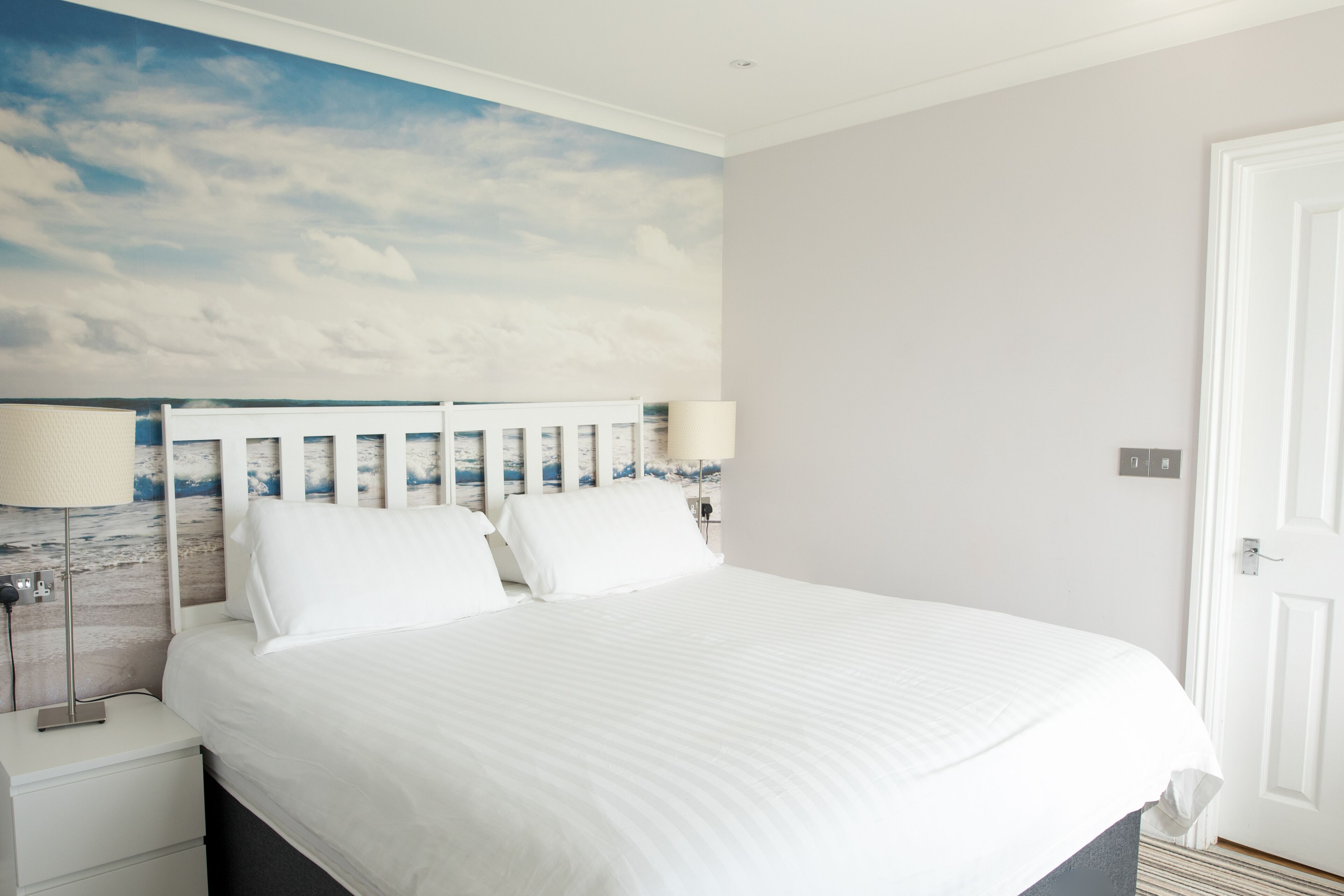 Cama bem arrumada com lençóis brancos, cabeceira com céu e nuvens pintadas e mesa de cabeceira branca com abajur.