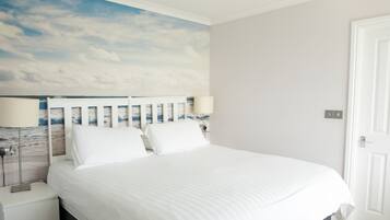 Cama bem arrumada com lençóis brancos, cabeceira com céu e nuvens pintadas e mesa de cabeceira branca com abajur.