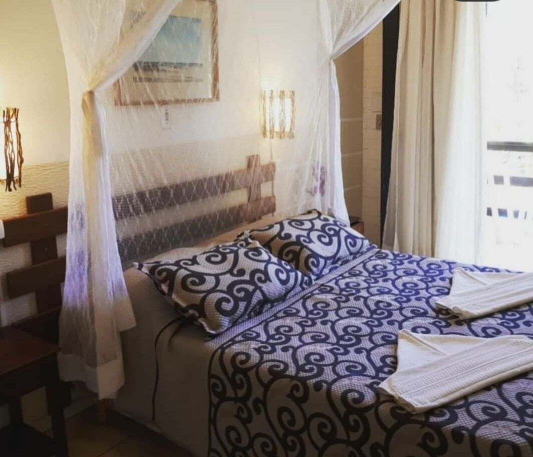 Uma cama com colcha estampada, mesa de cabeceira, uma cadeira e uma janela com cortinas.