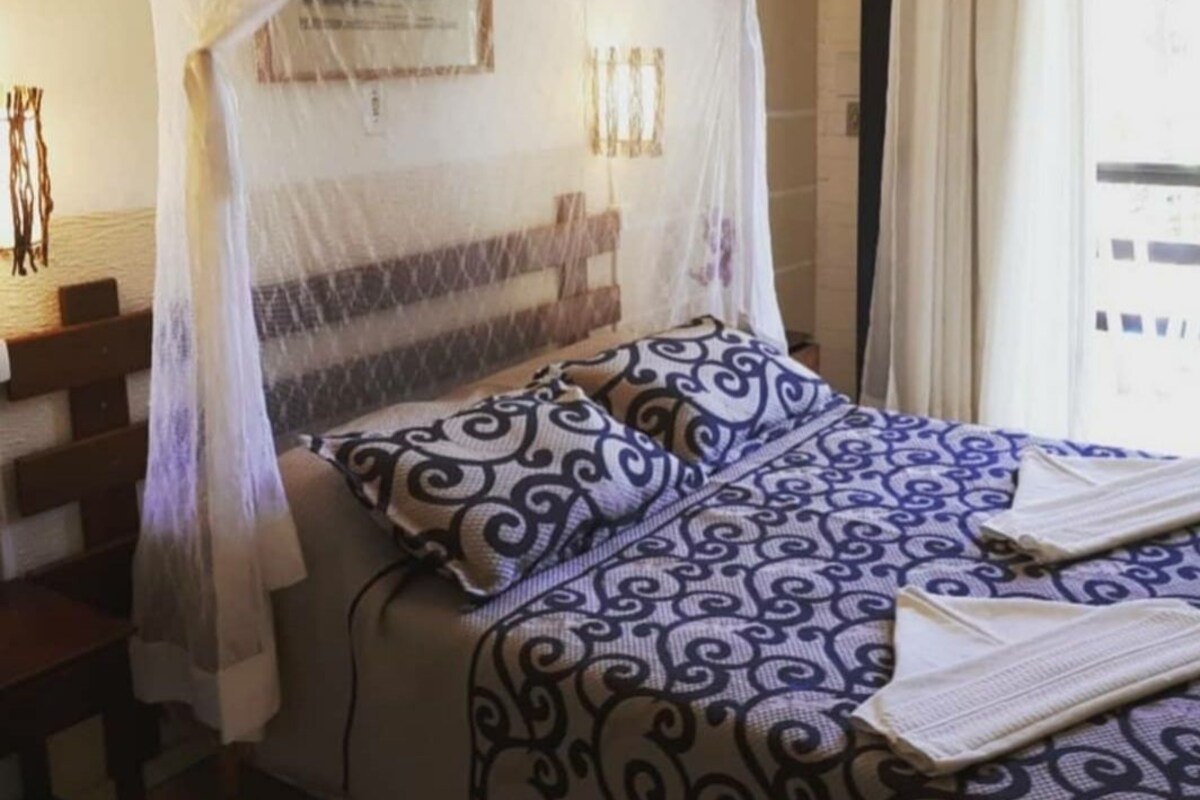 Uma cama com colcha estampada, mesa de cabeceira, uma cadeira e uma janela com cortinas.