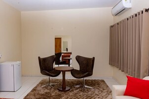 Uma sala de estar moderna com um sofá branco, duas poltronas, uma mesa redonda pequena e um travesseiro vermelho. Há um ar condicionado branco e um radiador com cortinas.