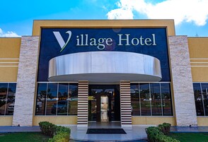 Exterior do Village Hotel, com entrada notável, janelas amplas e céu claro.