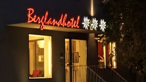 Entrada de um hotel com um letreiro de neon escrito "Berglandhotel" e uma escadaria que leva à porta.