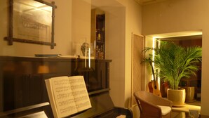 Um piano de cauda com partituras, uma poltrona de vime, um violão e uma planta em vaso num quarto com iluminação acolhedora.