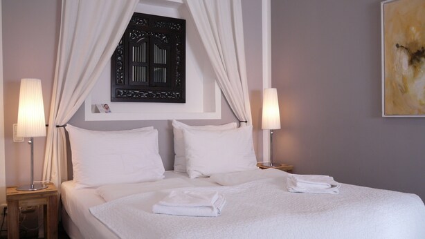 Cama bem arrumada com lençóis brancos e toalha dobrada, duas luminárias de apoio e um objeto decorativo na parede acima da cama.