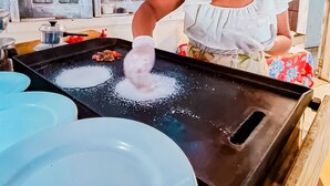 Uma pessoa cozinha em um fogão grande ao ar livre, com uma janela e uma porta ao fundo.