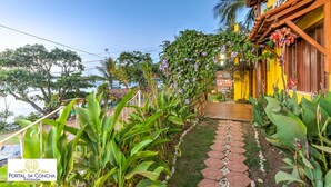 Uma trilha leva a um edifício com fachada amarela, cercado por vegetação exuberante e uma placa com a inscrição 'Portal da Concha Pousada'.