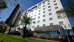 Um hotel moderno com vários andares, grandes janelas e um jardim bem cuidado com palmeiras e flores.