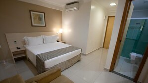 Quarto de hotel com cama, mesas de cabeceira, sofá, ar condicionado e banheiro visível através de uma porta de vidro.