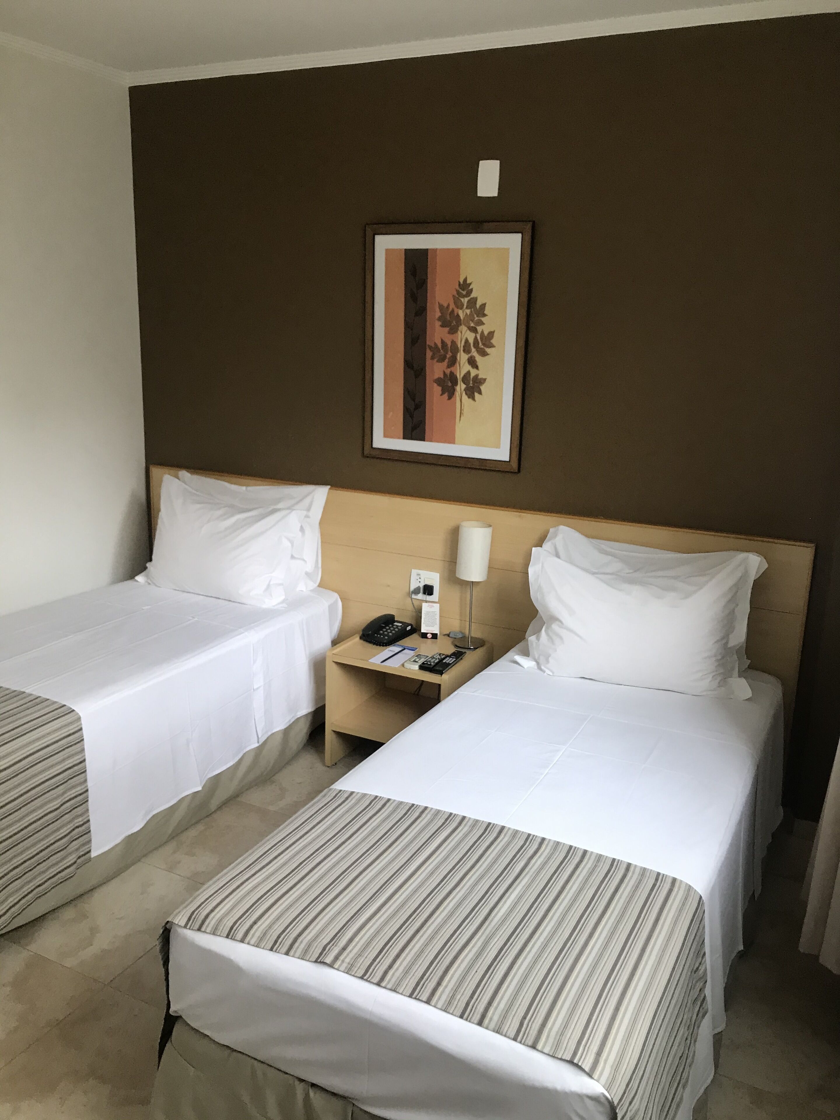 Quarto de hotel com duas camas de solteiro, mesa de cabeceira com telefone e abajur, e quadro na parede.
