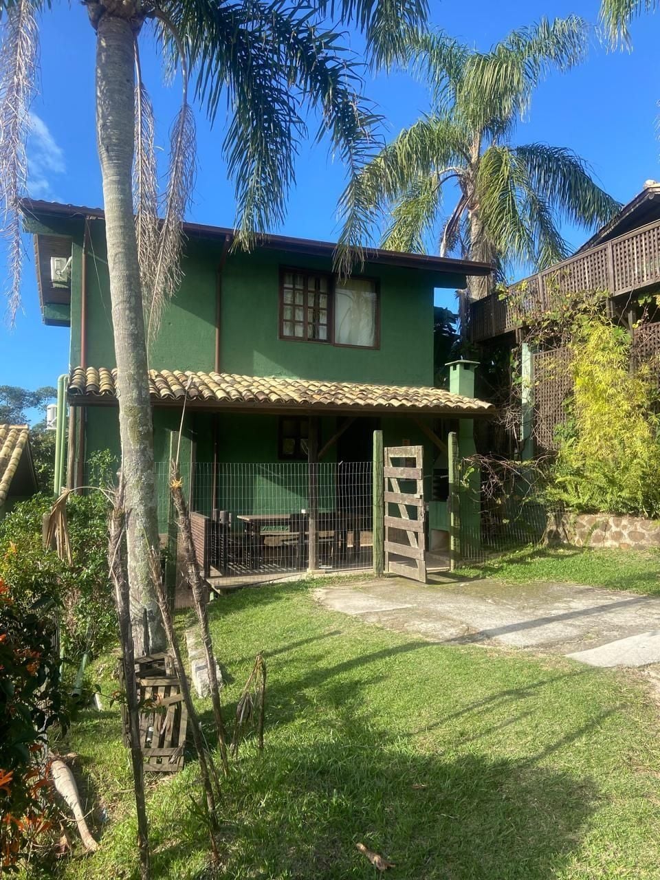 Uma casa verde com telhado de telhas, um quintal cercado e palmeiras.