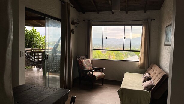 Quarto com cama, cadeira, mesa e vista para uma paisagem montanhosa através de grandes janelas.
