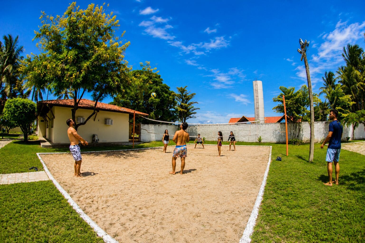 Uma quadra de vôlei de areia com jogadores, cercada por vegetação e um edifício com telhado vermelho.