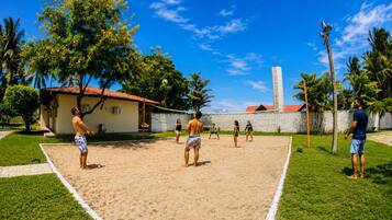 Uma quadra de vôlei de areia com jogadores, cercada por vegetação e um edifício com telhado vermelho.