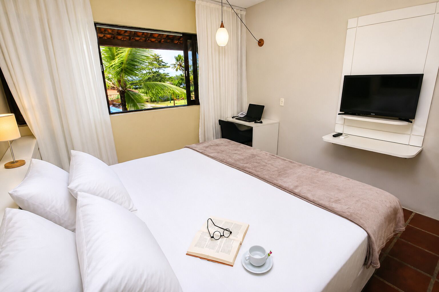 Uma cama bem arrumada com lençóis brancos, uma mesa de cabeceira com abajur, uma TV na parede e uma janela com vista para palmeiras.