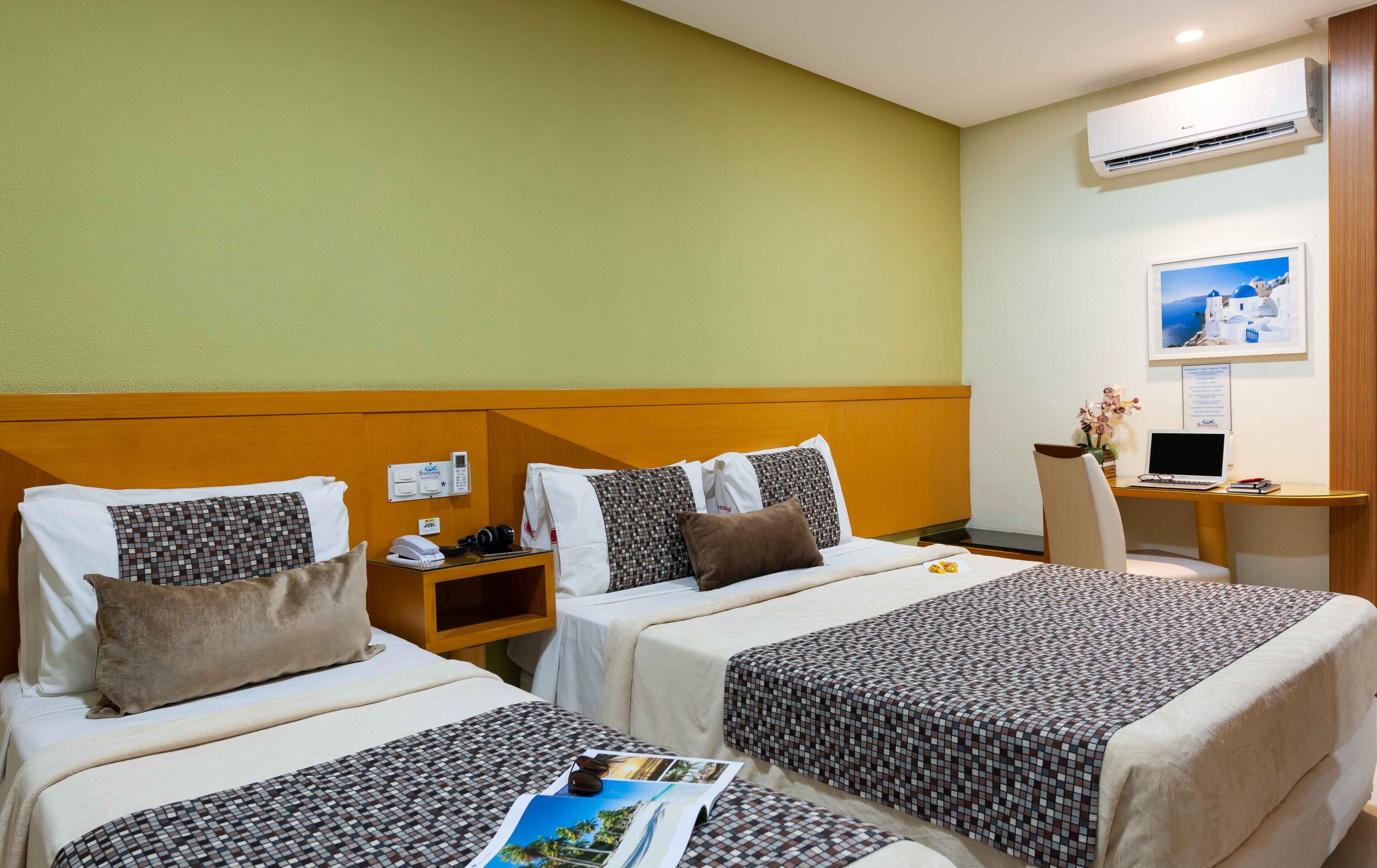 Quarto de hotel com duas camas, uma escrivaninha, uma cadeira, ar condicionado e um quadro na parede.