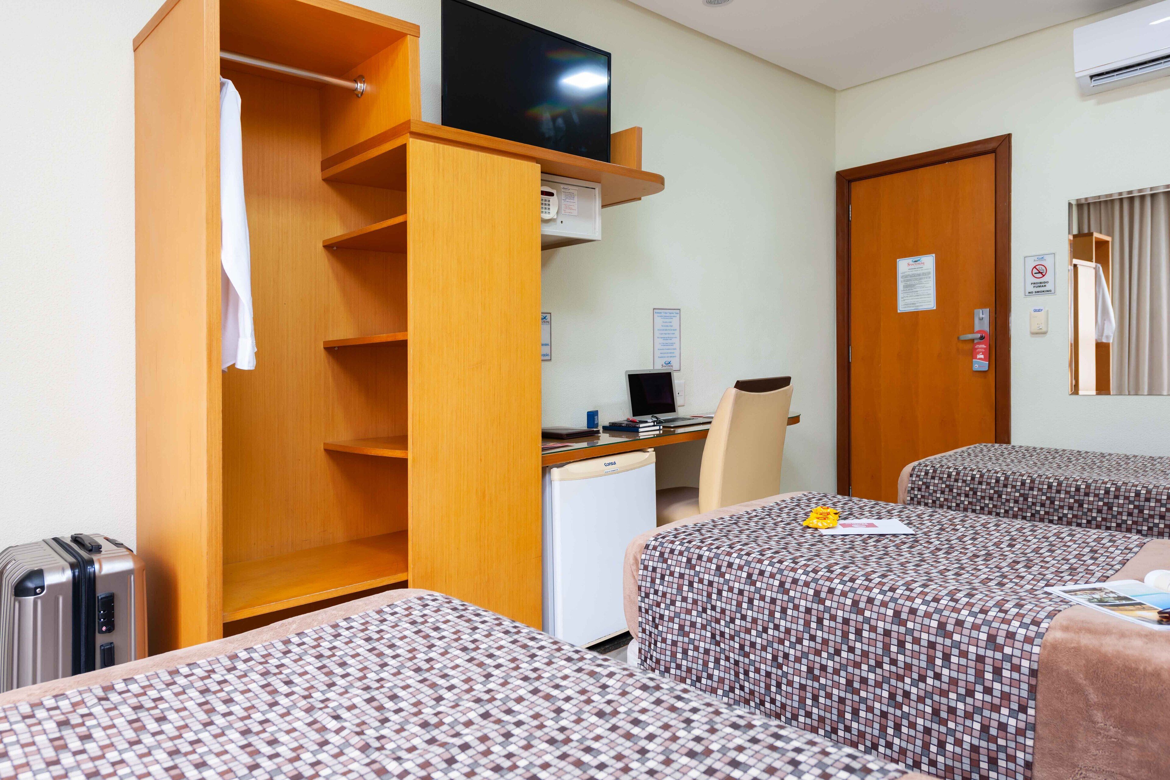 Quarto de hotel com duas camas, uma escrivaninha, televisão, guarda-roupa e um pequeno frigorífico.