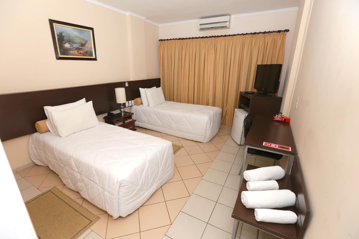 Quarto de hotel com duas camas, uma escrivaninha com computador, televisão e um quadro na parede.