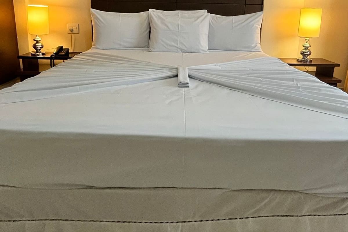 Uma cama grande com cabeceira escura, duas mesas de cabeceira com luminárias e uma colcha bem arrumada.