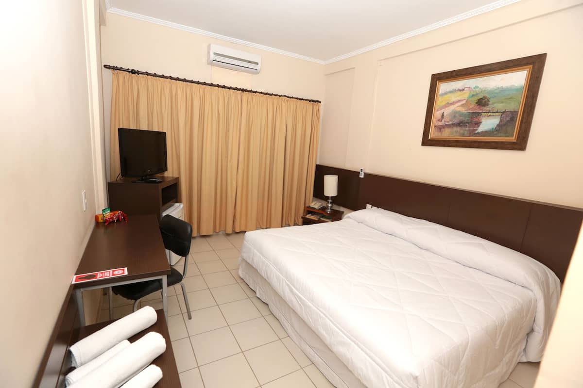 Quarto de hotel com cama, escrivaninha com televisão, uma cadeira, cortina e um quadro na parede, além de uma placa na escrivaninha.