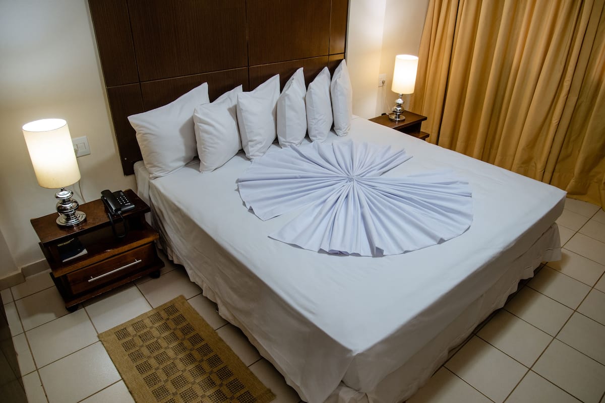 Cama bem arrumada com roupa de cama branca e arranjo decorativo de toalhas brancas. Tem uma mesa de cabeceira de madeira com uma luminária e um telefone. Um tapete bege está no chão.