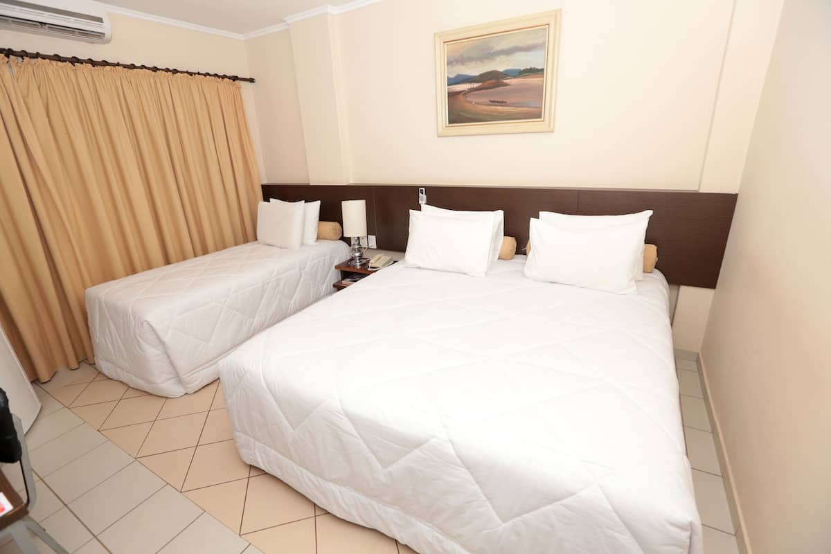 Quarto de hotel com duas camas, cortinas bege, um quadro na parede e piso em azulejo.