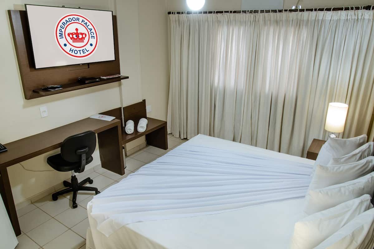 Quarto de hotel com cama, escrivaninha e cadeira, televisão na parede e janela com cortinas.