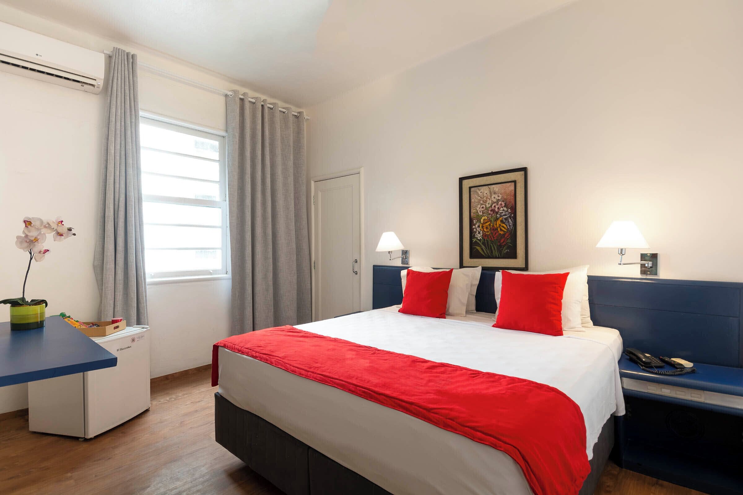 Quarto de hotel com cama, mesas de cabeceira, escrivaninha, cadeira, janela com cortinas, ar condicionado fixado na parede e um quadro emoldurado.