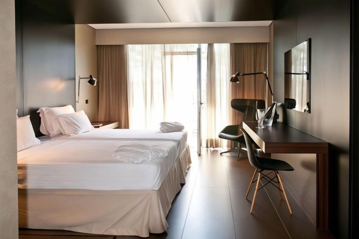 Quarto de hotel moderno com uma cama grande, uma escrivaninha e uma cadeira. Há uma janela com cortinas, um abajur e um espelho na parede.