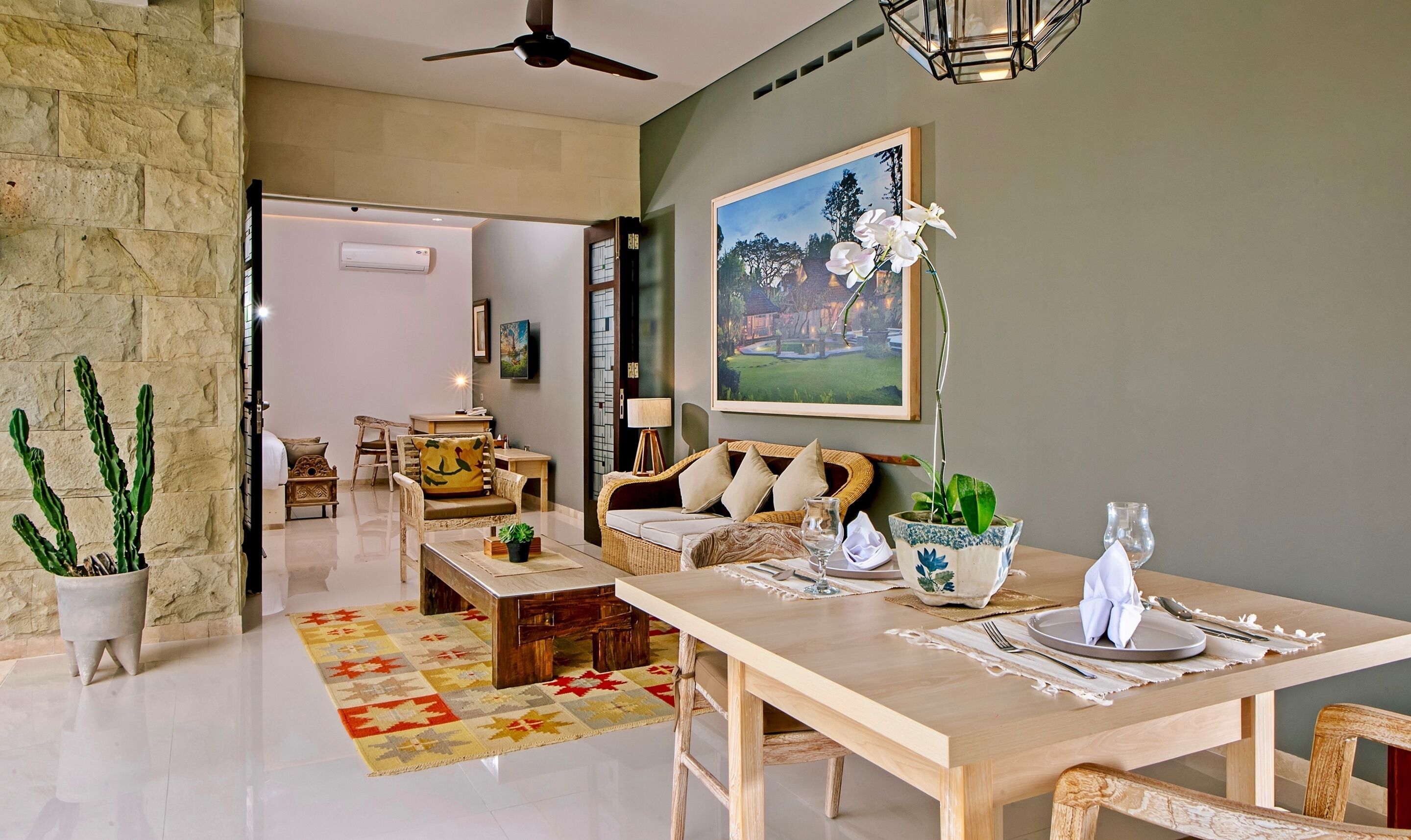 Ampla sala de estar com uma mesa de jantar posta para dois, um sofá e uma mesa de centro. Há uma pintura na parede e um ventilador de teto.
