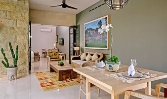 Ampla sala de estar com uma mesa de jantar posta para dois, um sofá e uma mesa de centro. Há uma pintura na parede e um ventilador de teto.