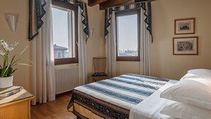 Quarto com cama, duas janelas com cortinas, radiador, cadeira e duas molduras na parede.