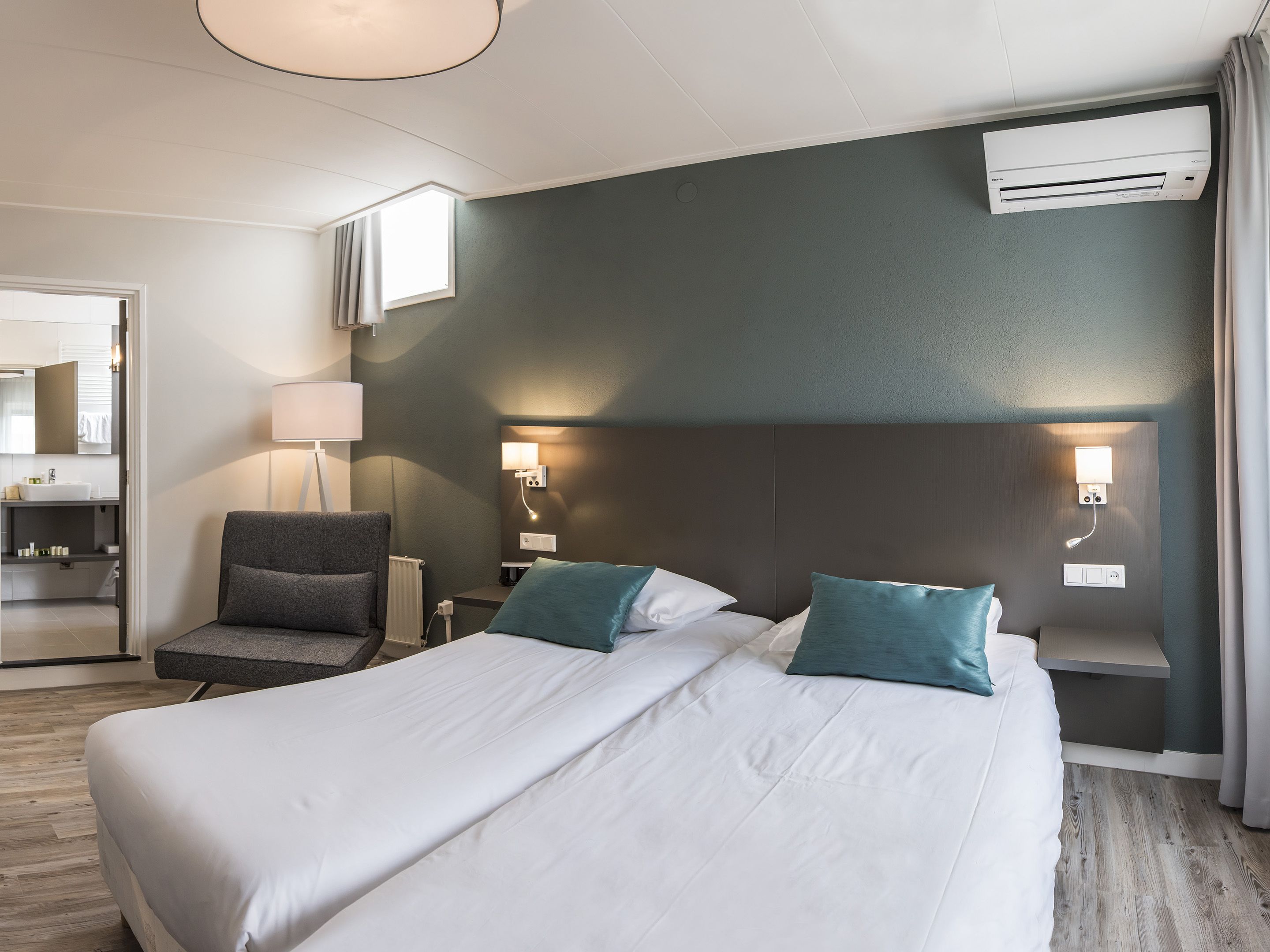 Quarto de hotel com uma cama grande, duas mesas de cabeceira, uma cadeira, uma escrivaninha, um espelho e um aparelho de ar condicionado.