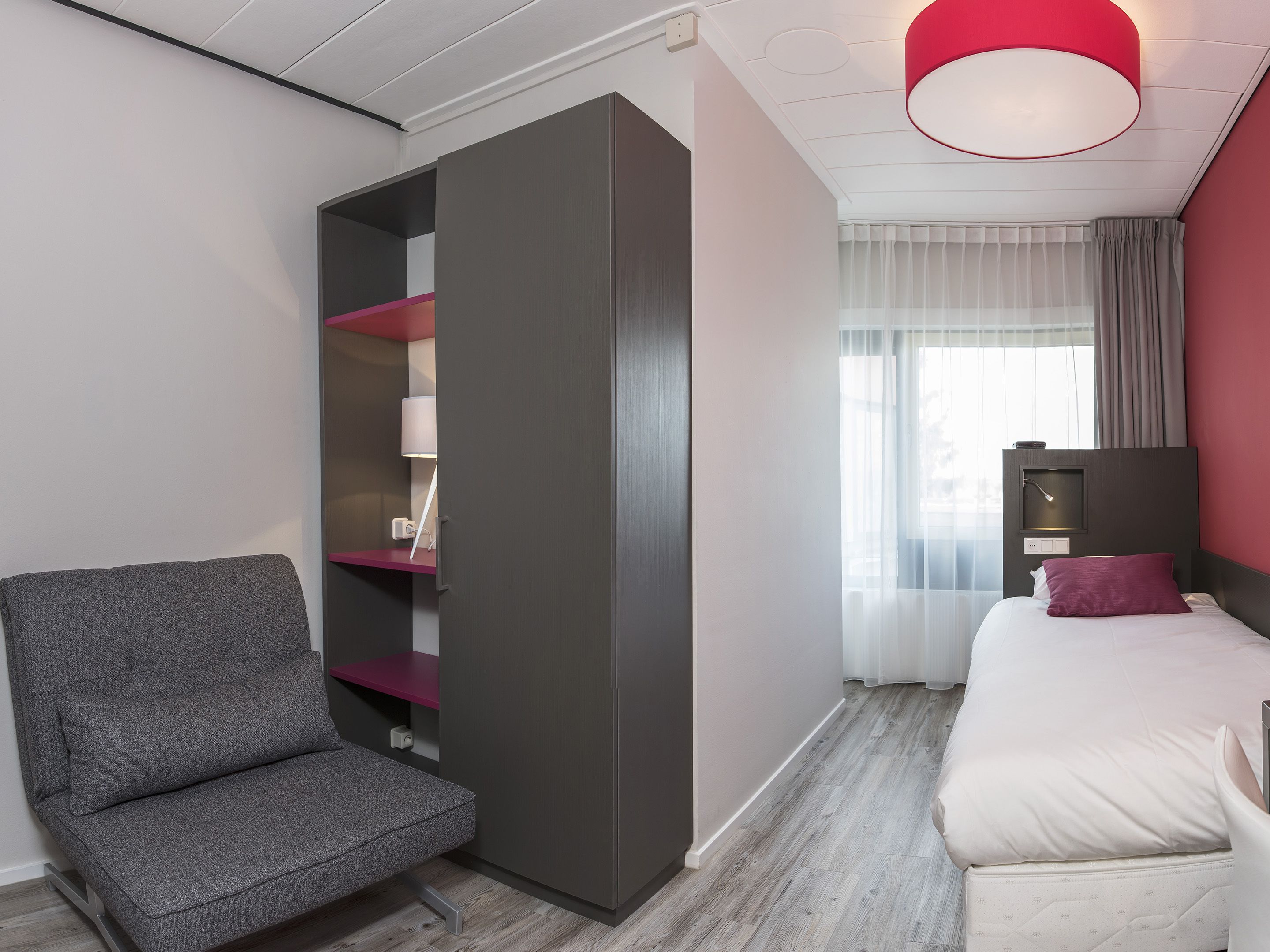 Quarto de hotel moderno com sofá cinzento, cama com colchão branco e almofada roxa, guarda-roupa cinzento com prateleira vermelha e uma luminária de teto vermelha.