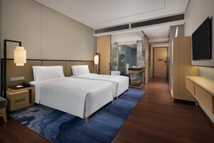 Quarto de hotel moderno com duas camas, uma escrivaninha, uma TV e um banheiro visível através de uma porta de vidro.