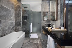 Uma casa de banho moderna com uma banheira independente, um chuveiro com proteção de vidro e uma toucador com bancada preta e painel de fundo em mármore.